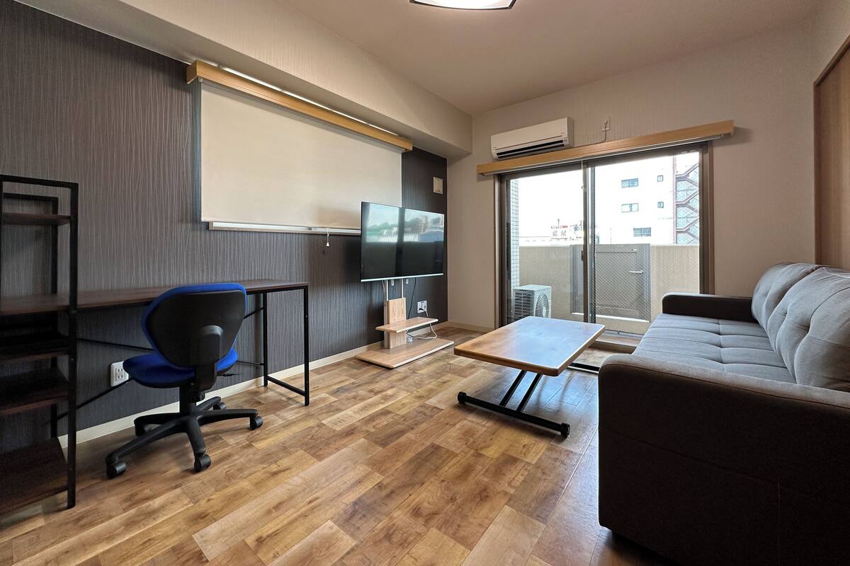 Uma sala de estar moderna com televisão de tela plana, um sofá, uma escrivaninha com cadeira e uma janela ampla com vista para edifícios.