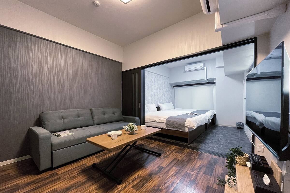 Quarto de hotel moderno com sofá cinza, mesa de centro de madeira, cama com cabeceira cinza e televisão em um suporte.