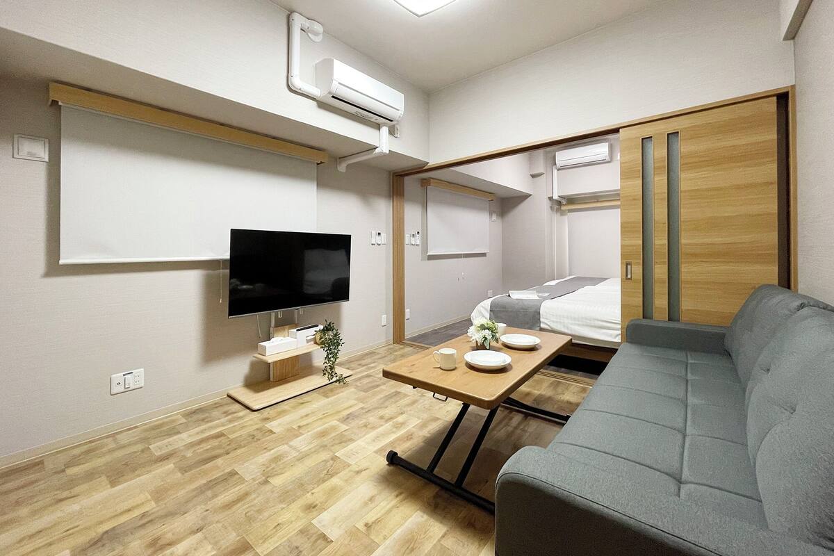 Quarto de hotel compacto com televisão de tela plana, um sofá, uma pequena mesa com uma planta e uma cama com cabeceira de madeira.