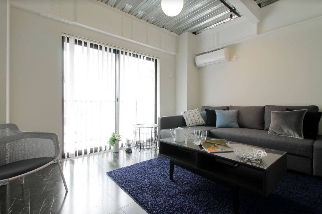 Uma sala de estar moderna com um sofá cinzento, uma mesa de centro e um tapete azul. Há uma poltrona e uma pequena mesa com uma planta.
