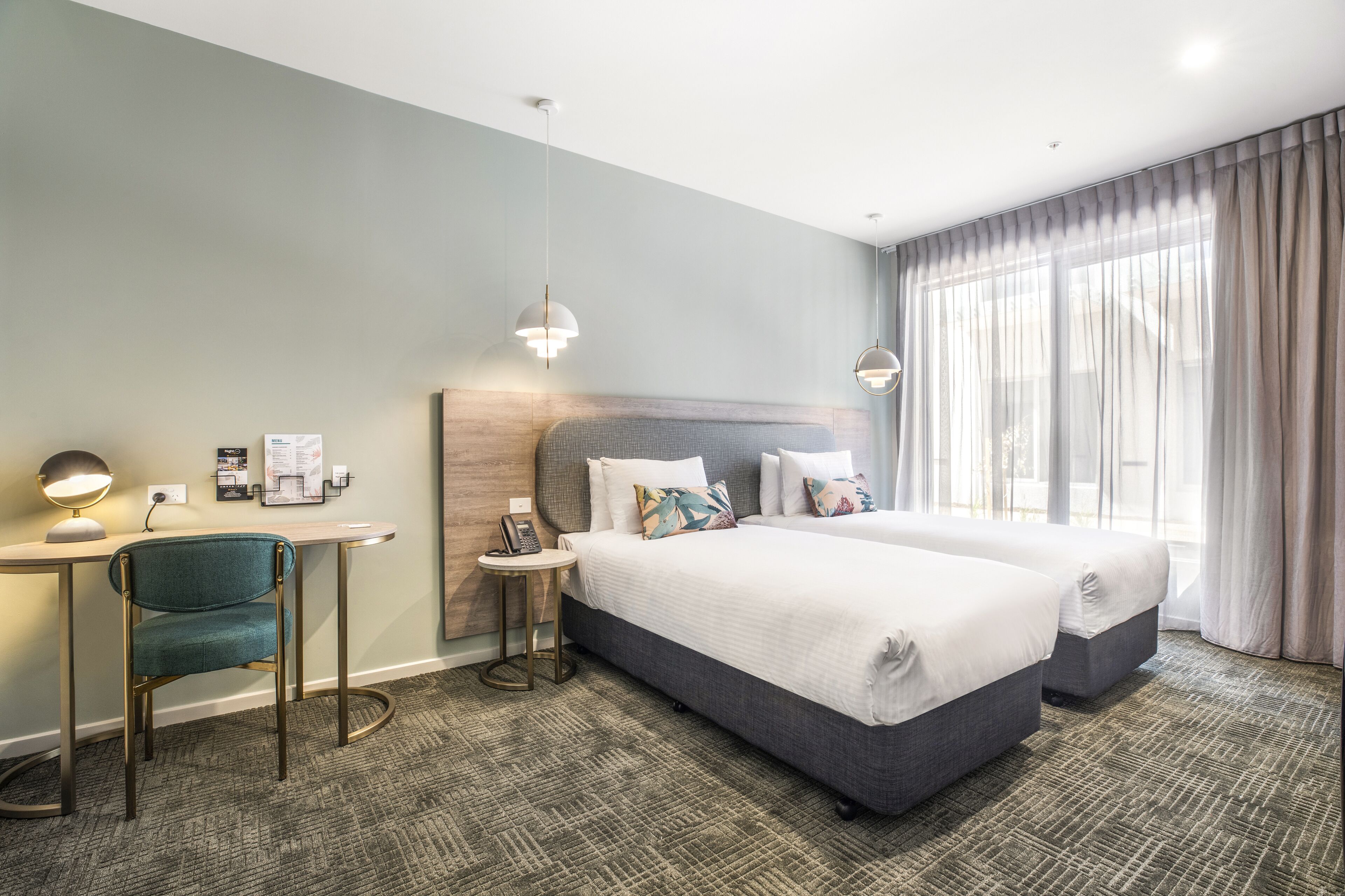 Quarto de hotel com duas camas, uma escrivaninha com cadeira, uma mesinha e um telefone fixado na parede.