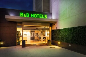 Exterior do B&B Hotels com design moderno, incluindo uma área de recepção e um conjunto de sebe verde.