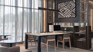 Quarto de hotel moderno com uma escrivaninha grande, duas cadeiras, um computador e uma janela ampla com cortinas leves.