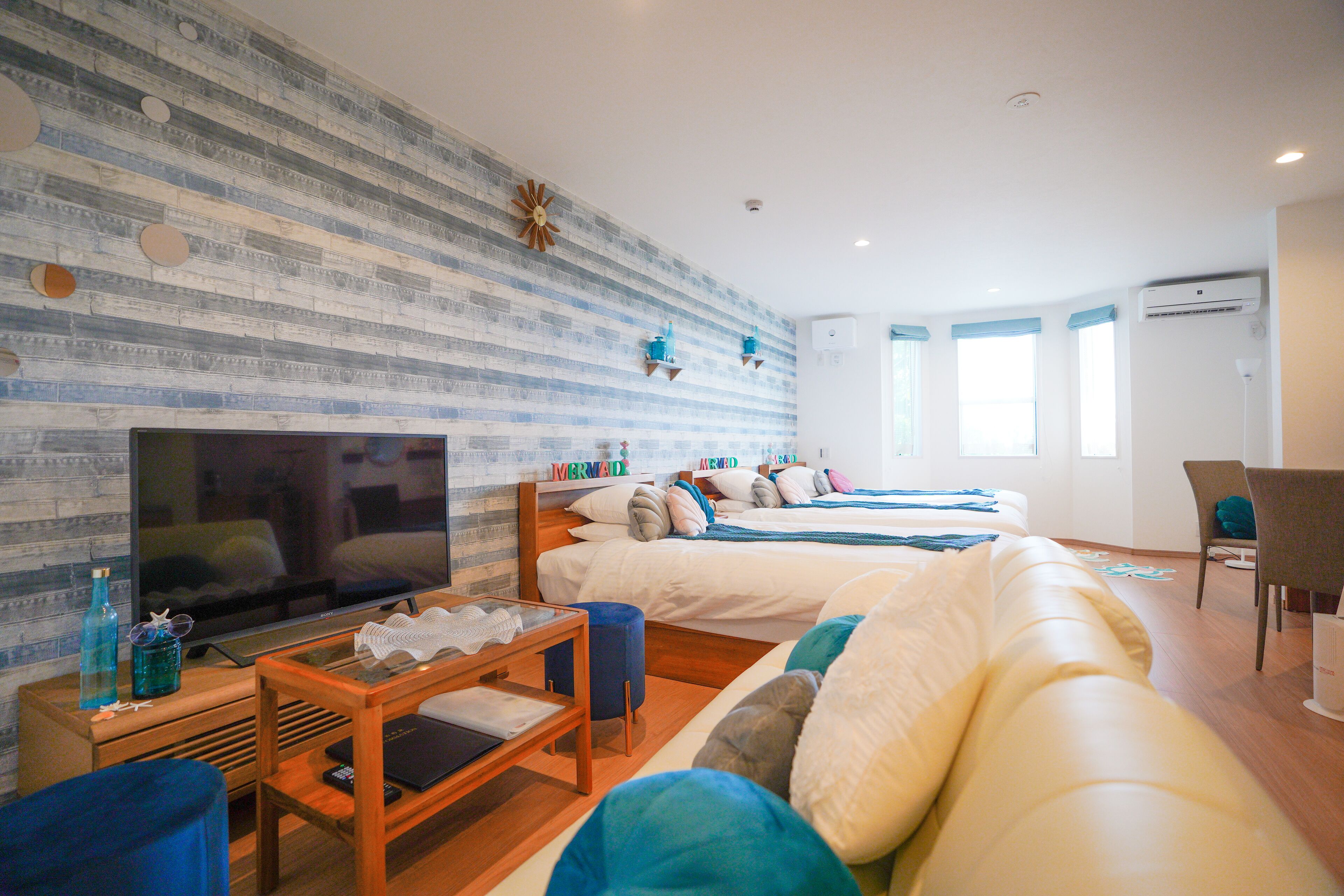 Quarto de hotel moderno com uma grande televisão de tela plana, um sofá, uma mesa de centro de madeira e uma cama com várias almofadas e um cobertor azul.