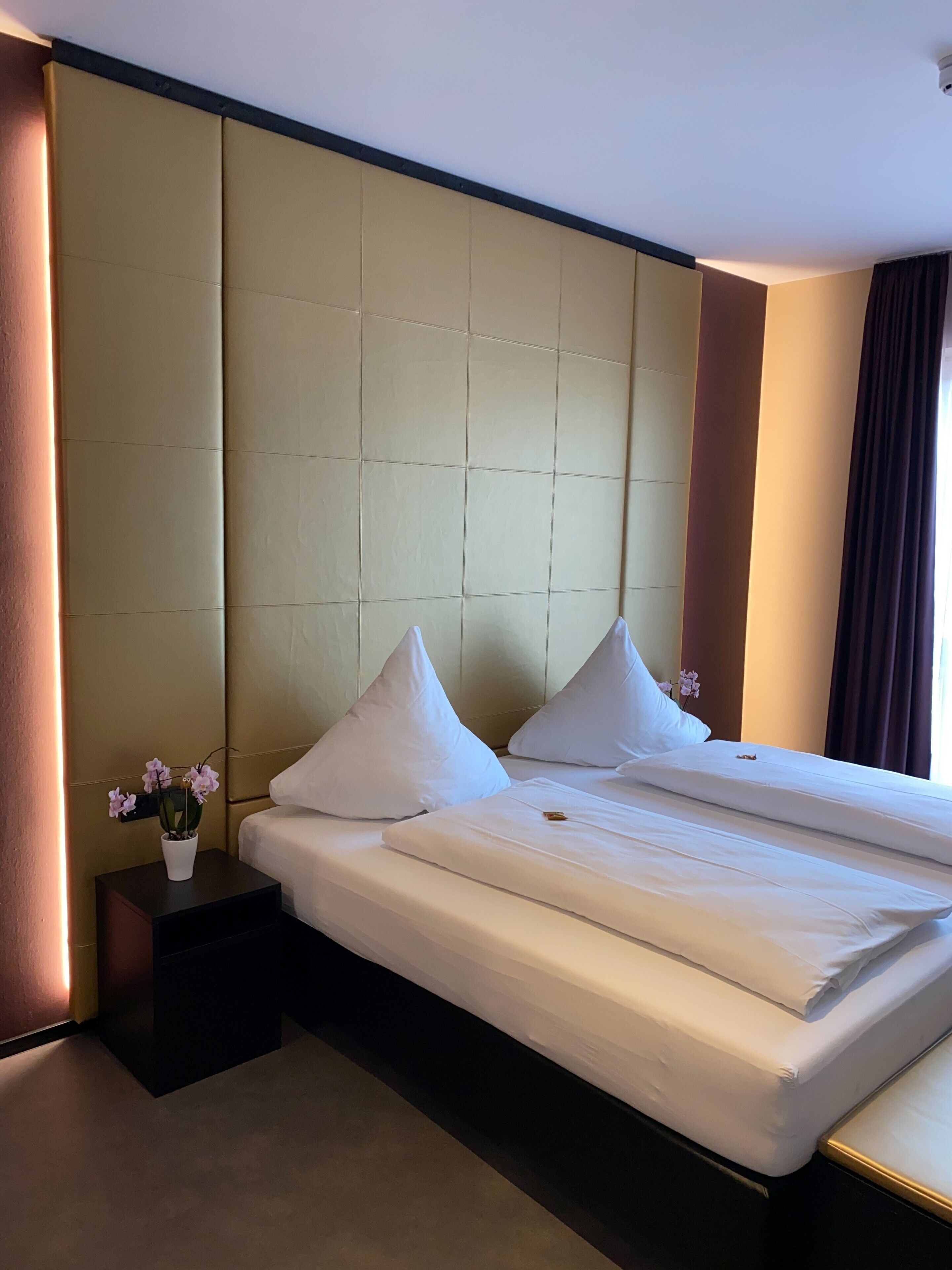 Quarto de hotel com uma cama grande, duas almofadas, mesa de cabeceira com um vaso de flores e cabeceira com painel de luz.