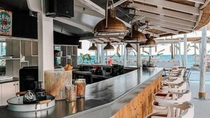 Uma área de bar com balcão de madeira, banquetas e luminárias pendentes.