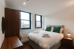 Quarto moderno com uma cama grande, duas janelas, uma escrivaninha e uma mesinha de cabeceira.