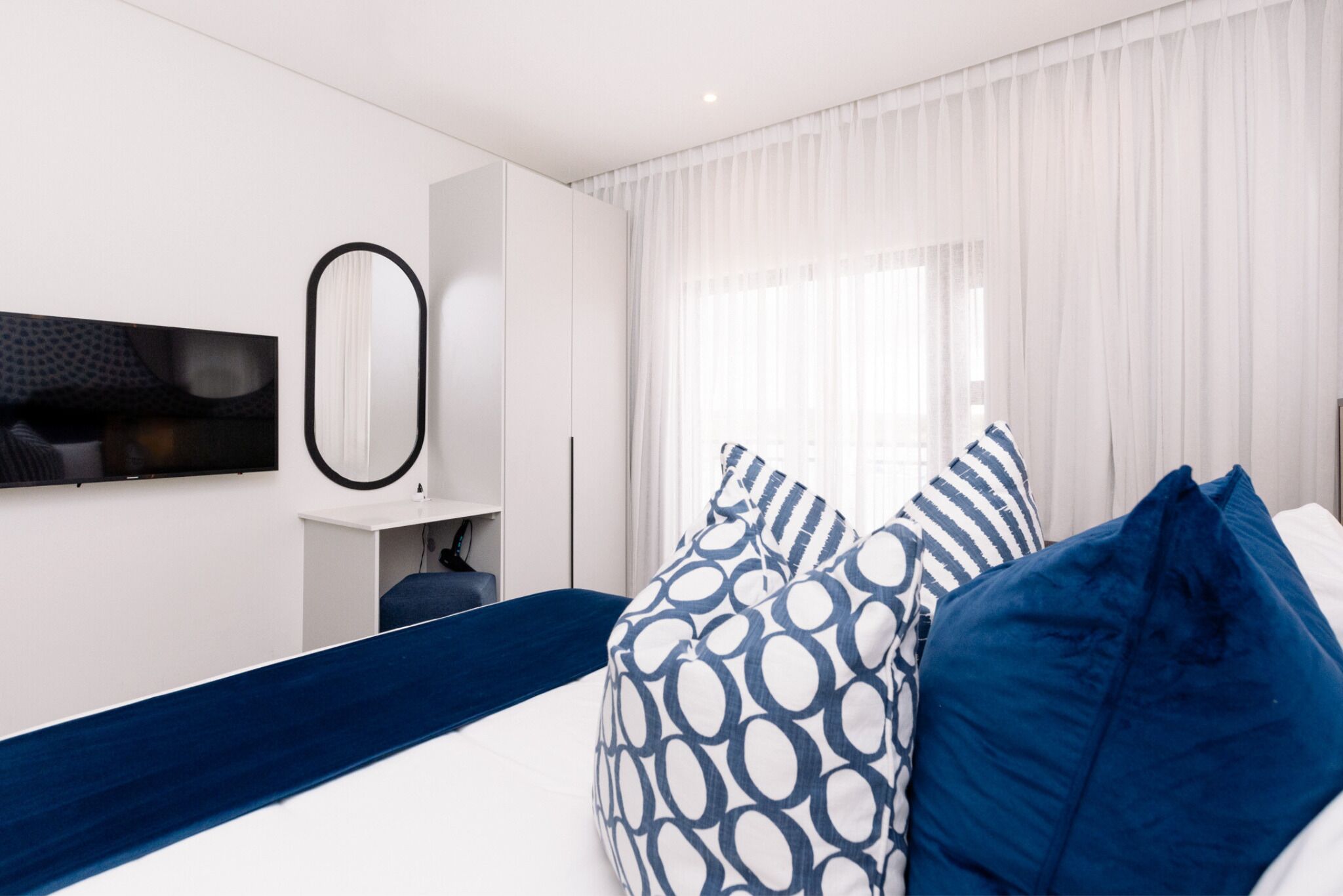 Quarto de hotel moderno com cama bem arrumada, um espelho grande e uma janela com cortinas leves.