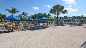 Área de piscina com guarda-sóis azuis, espreguiçadeiras e um calçadão pavimentado.