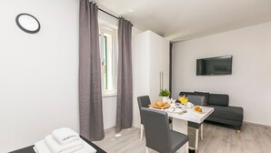 Um quarto moderno e bem iluminado, com uma mesa de jantar, cadeiras, um sofá, uma TV de tela plana e uma janela com cortinas.