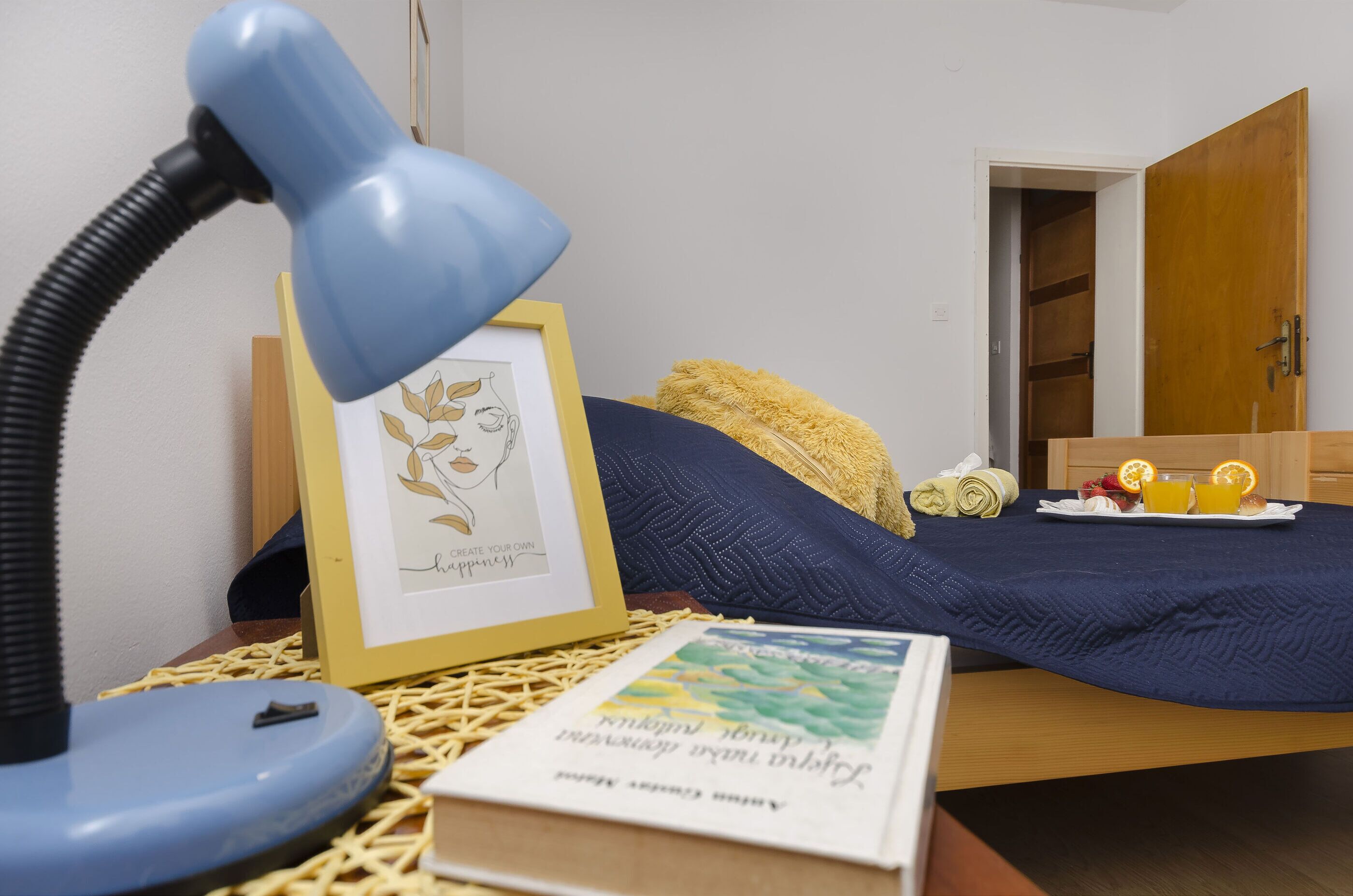 Cama bem arrumada com manta azul, quadro emoldurado, livro, abajur azul e bandeja com fatias de laranja e bebida sobre mesa de cabeceira de madeira.