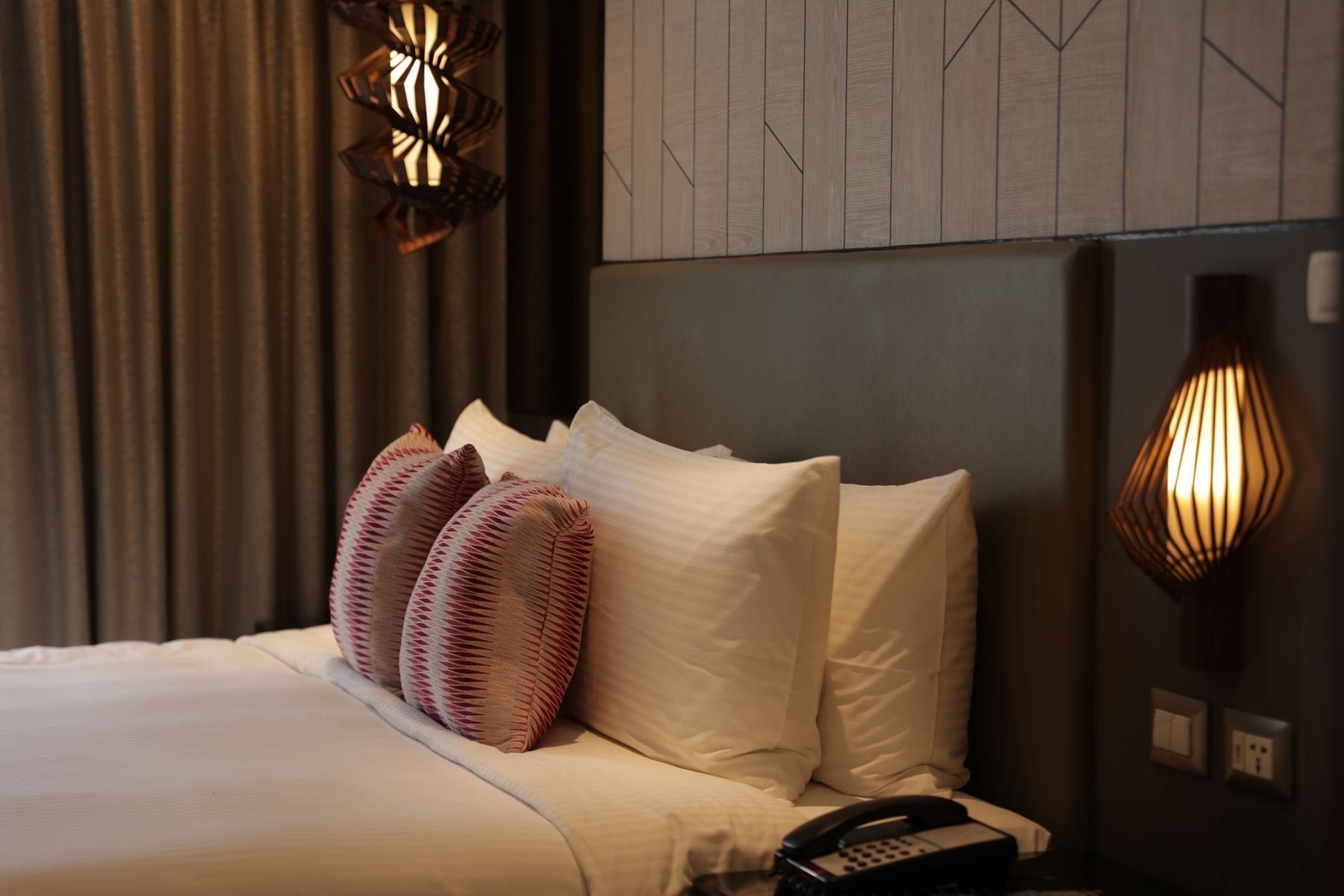 Quarto de hotel com cama, almofadas, telefone ao lado da cama e luminária na parede.