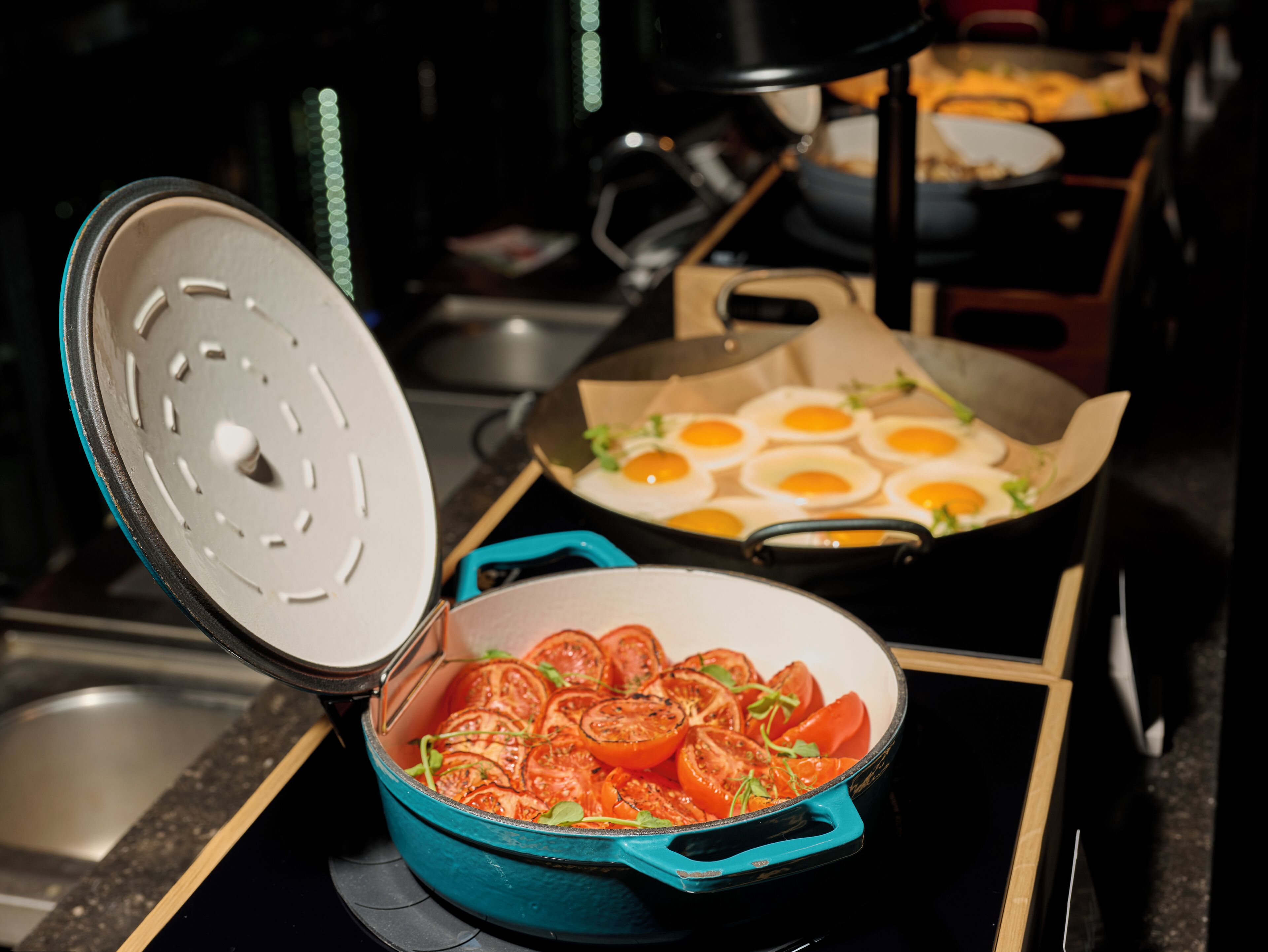 Uma cozinha com uma panela azul cheia de tomates fatiados em um fogão, uma frigideira com ovos e uma tampa de panela.