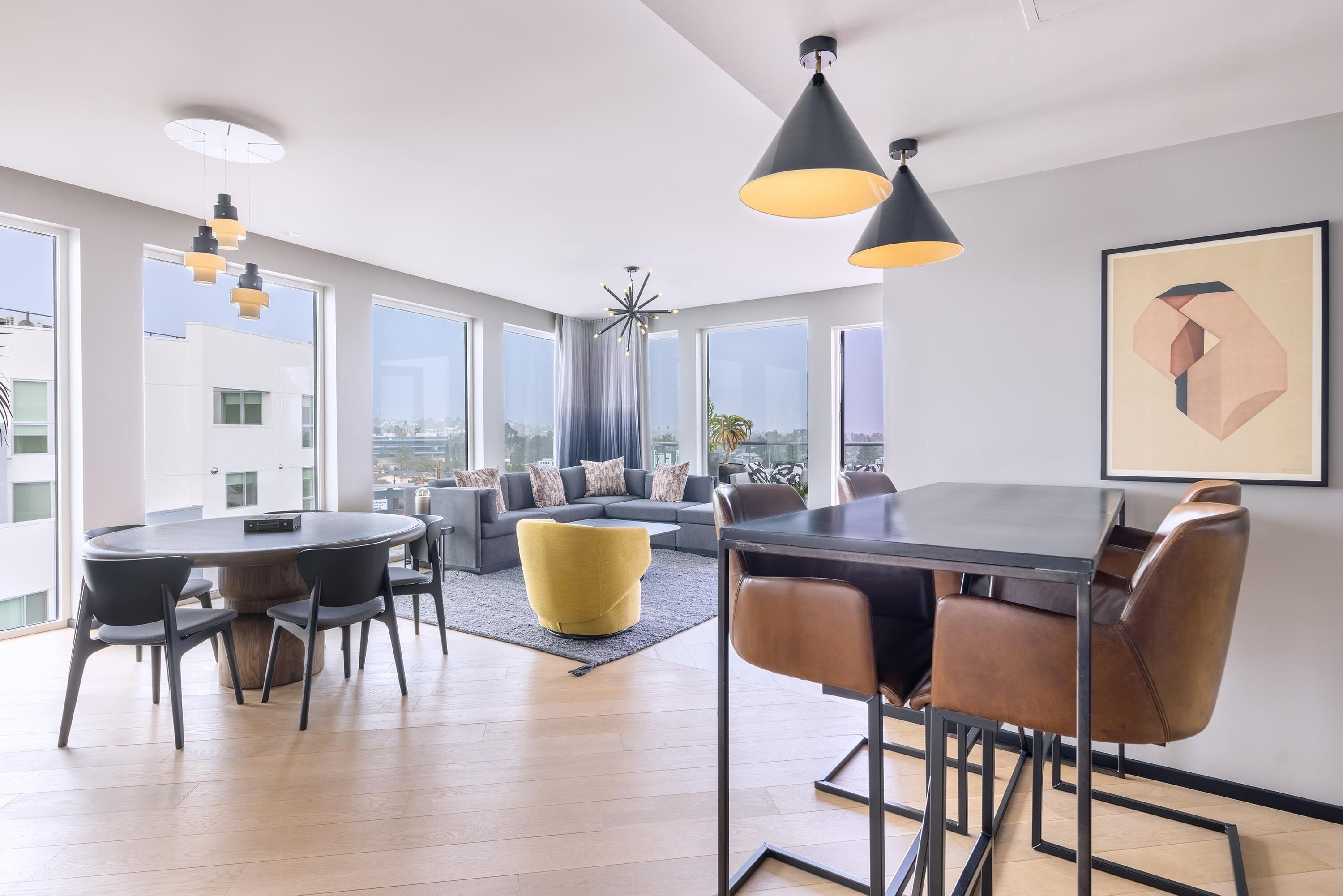 Um interior moderno com uma mesa de jantar redonda, cadeiras pretas, um banquinho amarelo, um sofá preto com almofadas e uma janela grande com vista para a cidade.