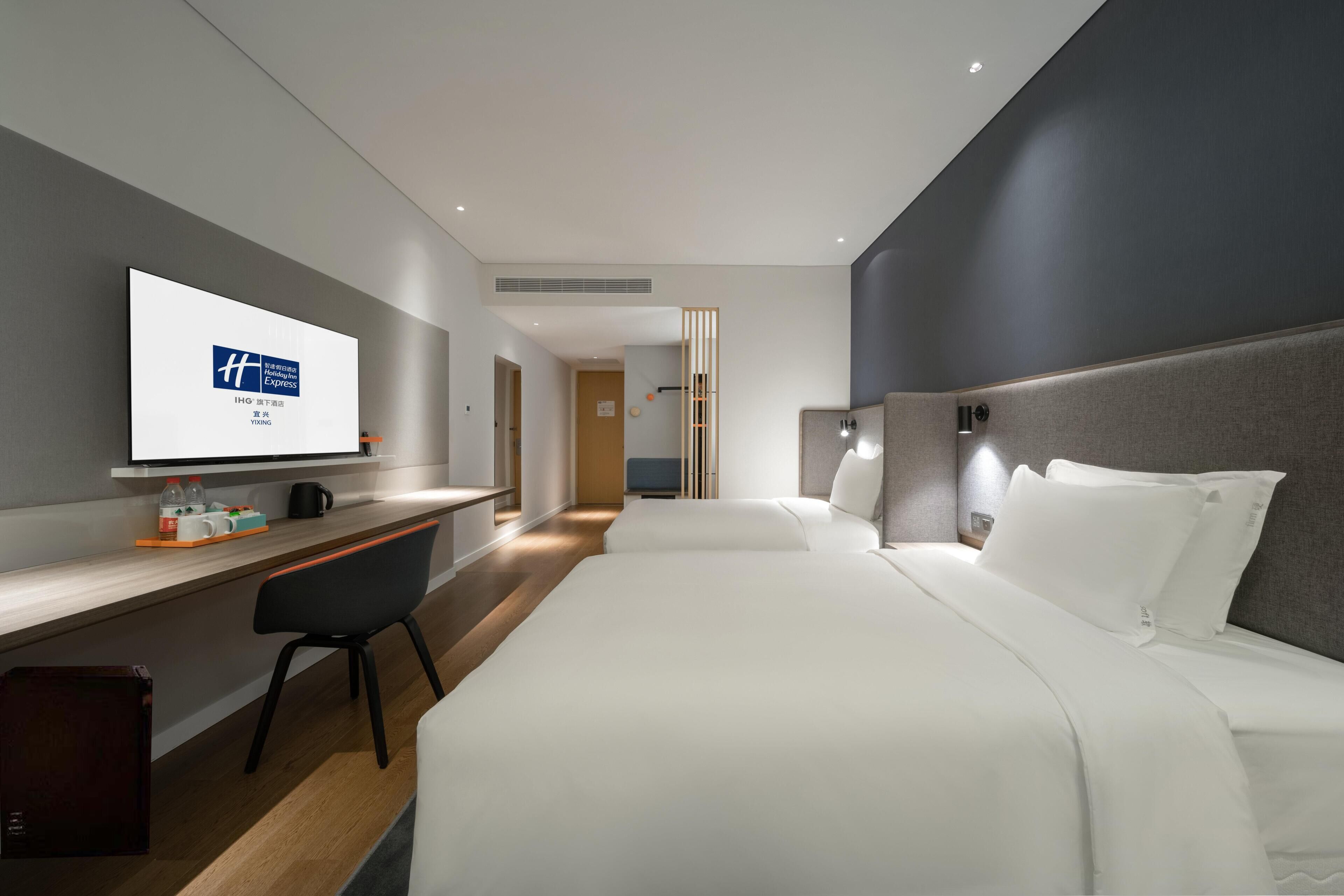 Quarto de hotel moderno com uma cama, uma escrivaninha e uma televisão exibindo o logotipo do hotel.