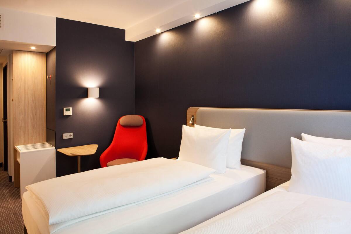 Quarto de hotel com uma cama grande, uma cadeira vermelha, uma mesa pequena e uma luminária fixada na parede.
