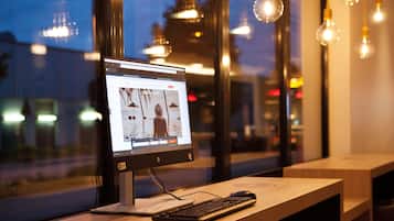 Um computador com monitor exibindo uma página web, conectado a um teclado e rato, colocado sobre uma mesa de madeira perto de uma janela com lâmpadas penduradas.