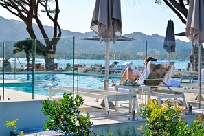 Uma área de piscina com espreguiçadeiras, guarda-sóis e uma barreira de vidro.