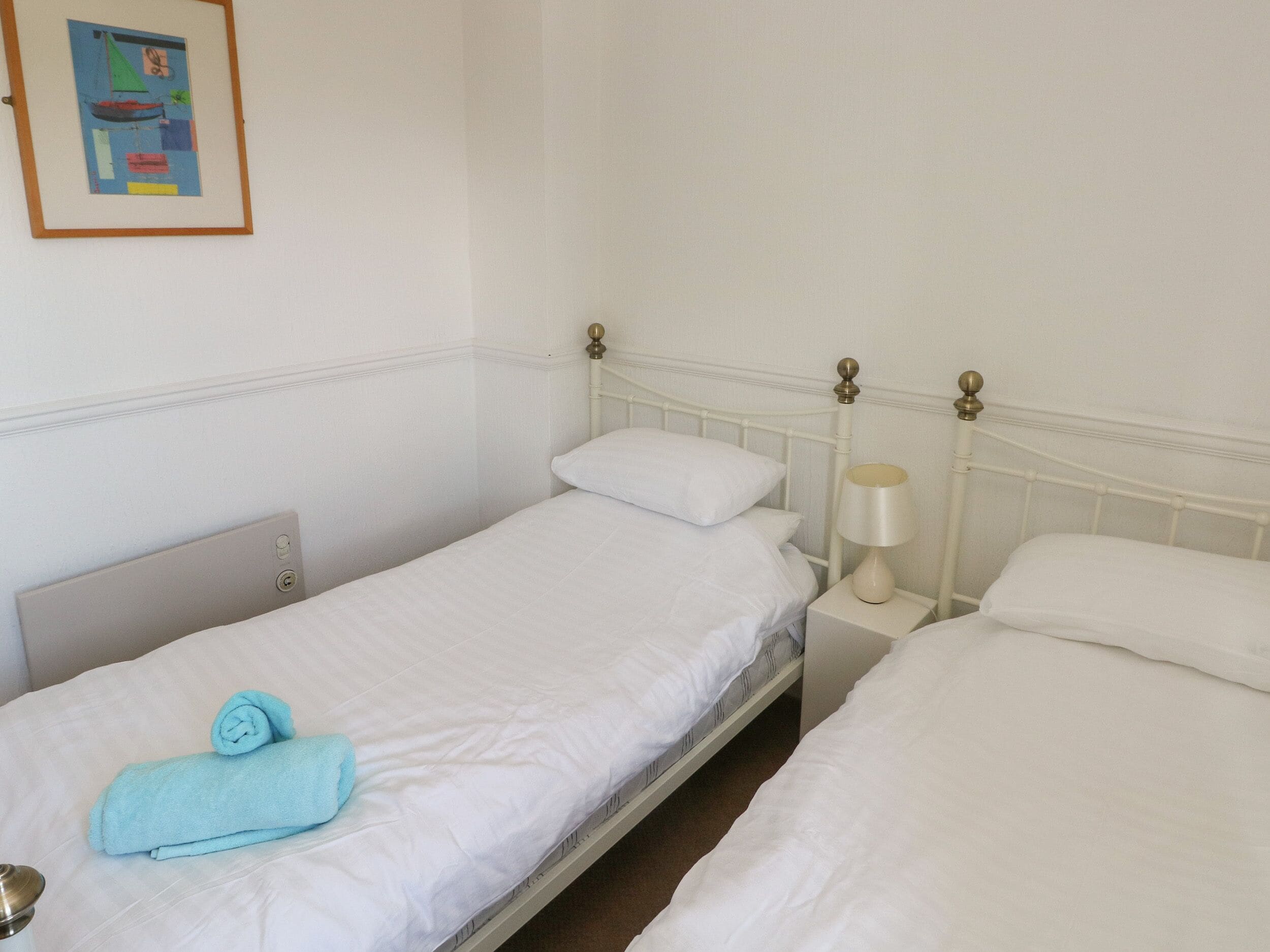 Quarto duplo com duas camas de solteiro, mesa de cabeceira com luminária, tomada na parede e um quadro na parede.