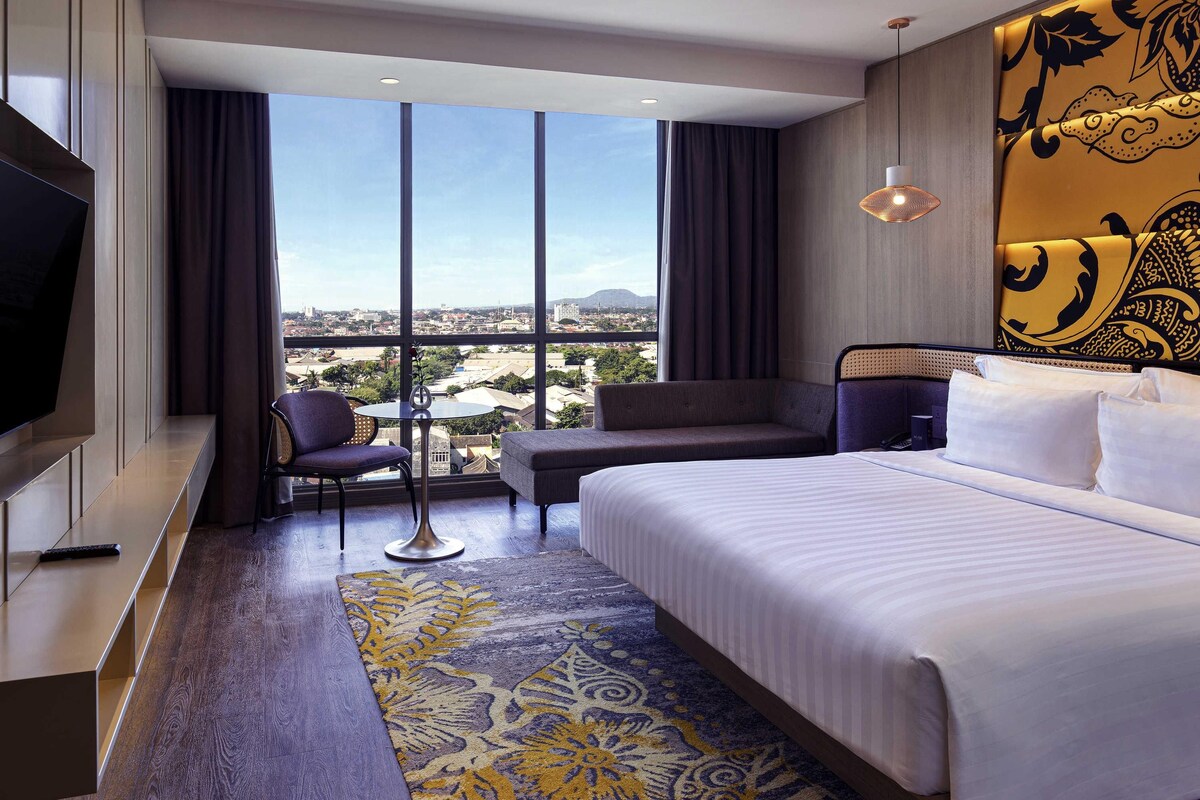Quarto de hotel com uma cama grande, área de estar, televisão e vista para a cidade.