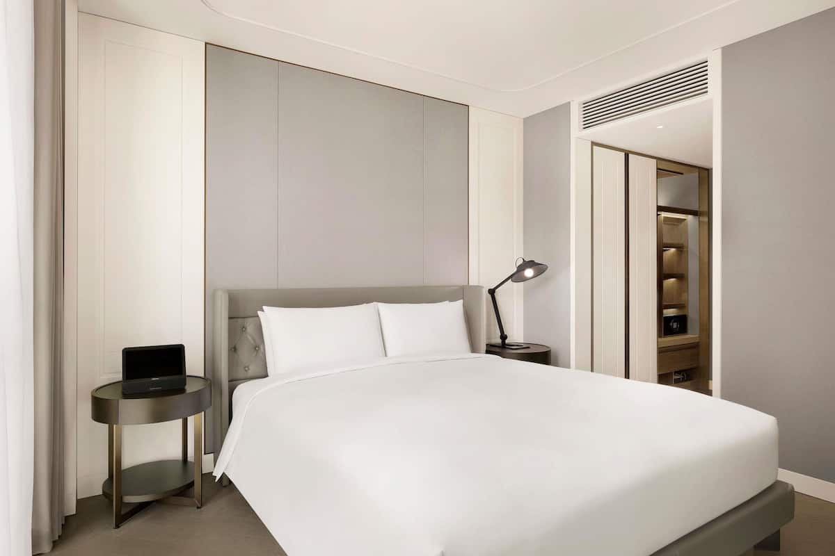 Quarto de hotel moderno com uma cama grande, mesa de cabeceira com abajur, guarda-roupa e espelho.