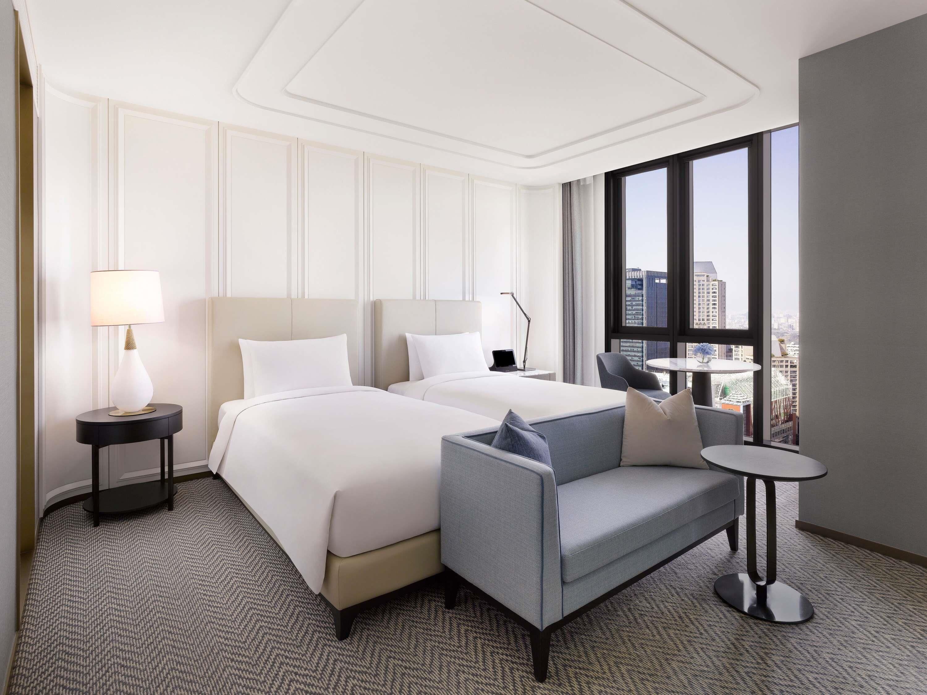 Quarto de hotel com duas camas, um sofá, uma pequena mesa e vista para a cidade.