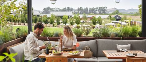 Uma área de refeições com uma grande janela que dá para um jardim e vista do campo. Há mesas e cadeiras de madeira, e um banco com almofadas.