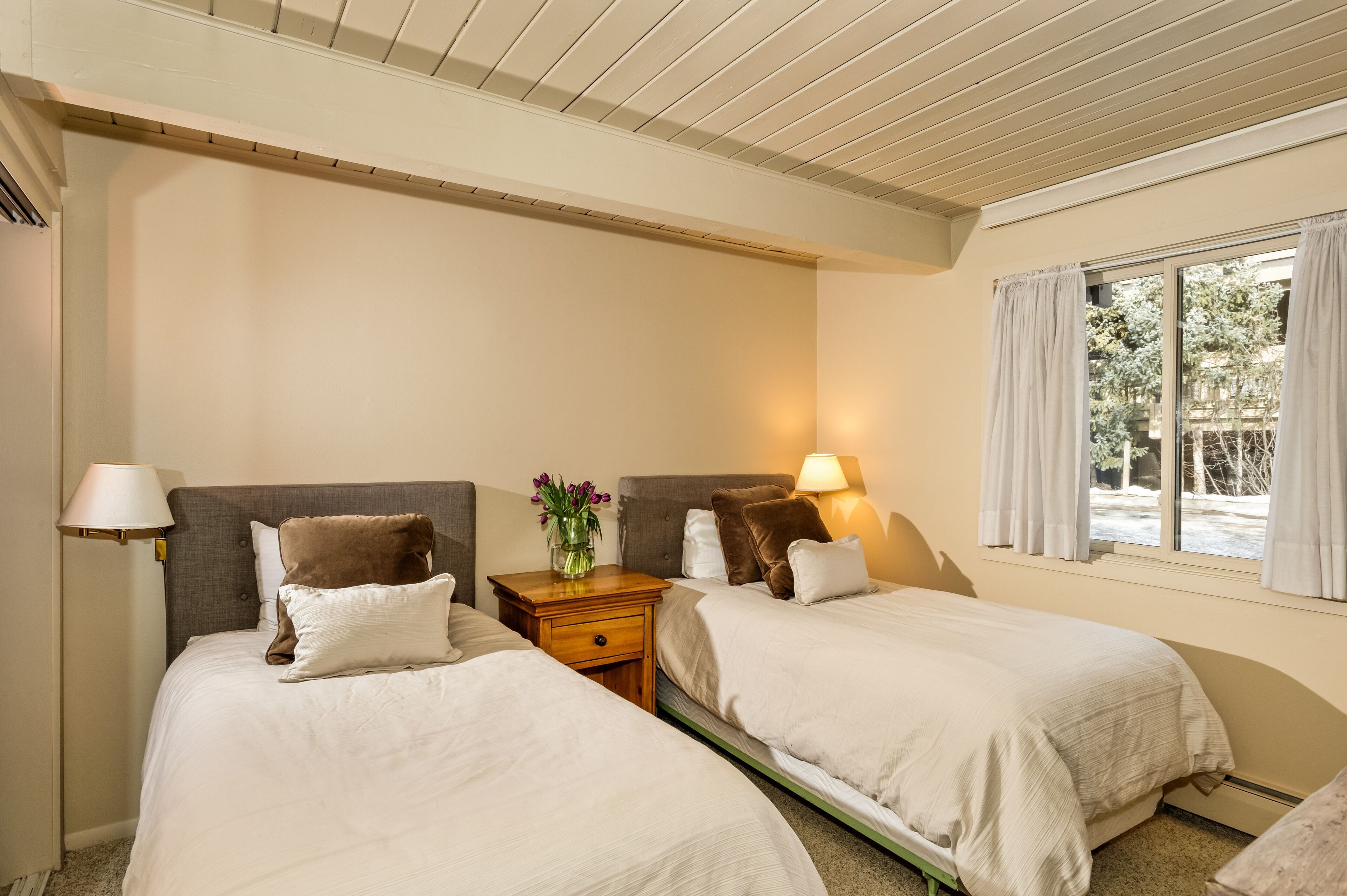 Quarto de hotel com duas camas, uma mesa de cabeceira com abajur e uma janela com cortinas.