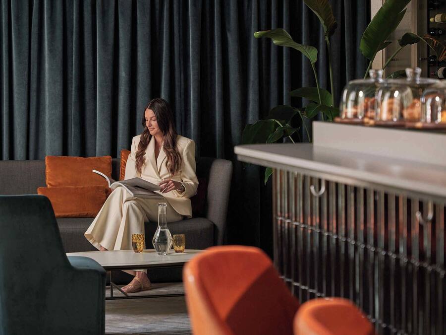 Mulher sentada num sofá numa sala moderna com espaço de bar e uma planta.