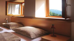 Quarto com cama, duas almofadas, mesa de cabeceira com abajur e janela com vista para montanhas.