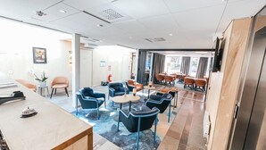 Um hall de hotel com balcão de recepção, área de estar com cadeiras e mesas, e vista para a sala de jantar.