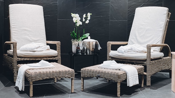 Um espaço de spa com duas cadeiras de vime, uma mesinha com um vaso de orquídeas brancas e uma parede revestida de azulejos pretos.