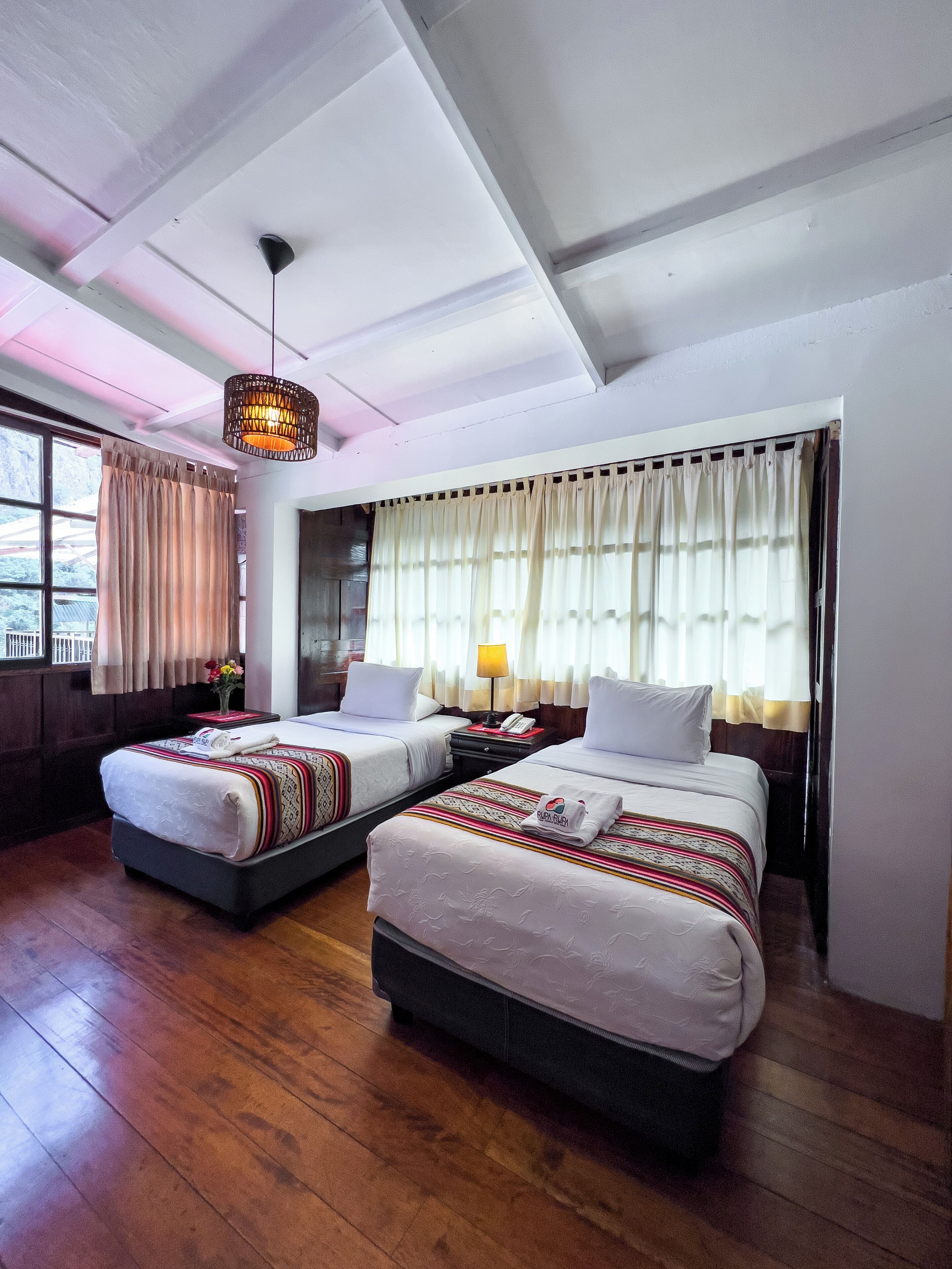 Quarto de hotel com duas camas, piso de madeira, luminária pendente e vista para o exterior.