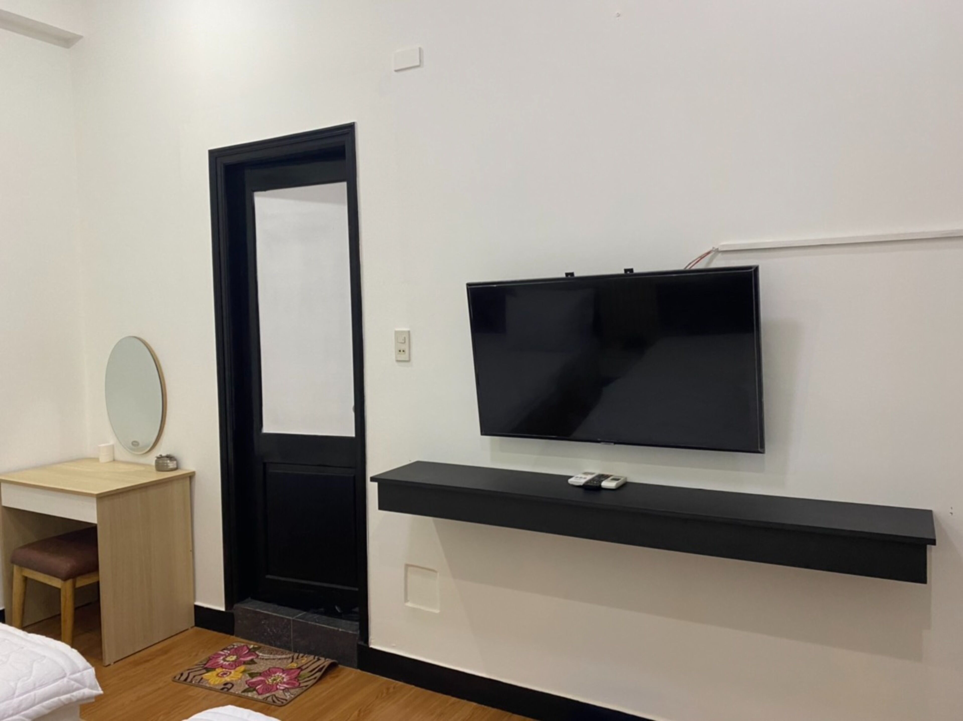 Quarto com cama, escrivaninha com cadeira, mesa pequena com espelho, televisão de tela plana presa em uma prateleira preta e uma porta.