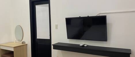 Quarto com cama, escrivaninha com cadeira, mesa pequena com espelho, televisão de tela plana presa em uma prateleira preta e uma porta.