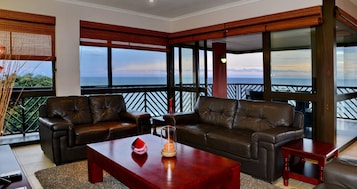 Uma sala de estar moderna com sofás de couro, uma mesa de centro de madeira e uma estrutura de vidro com uma vela. Grandes janelas oferecem vista para o oceano e uma varanda com corrimão.