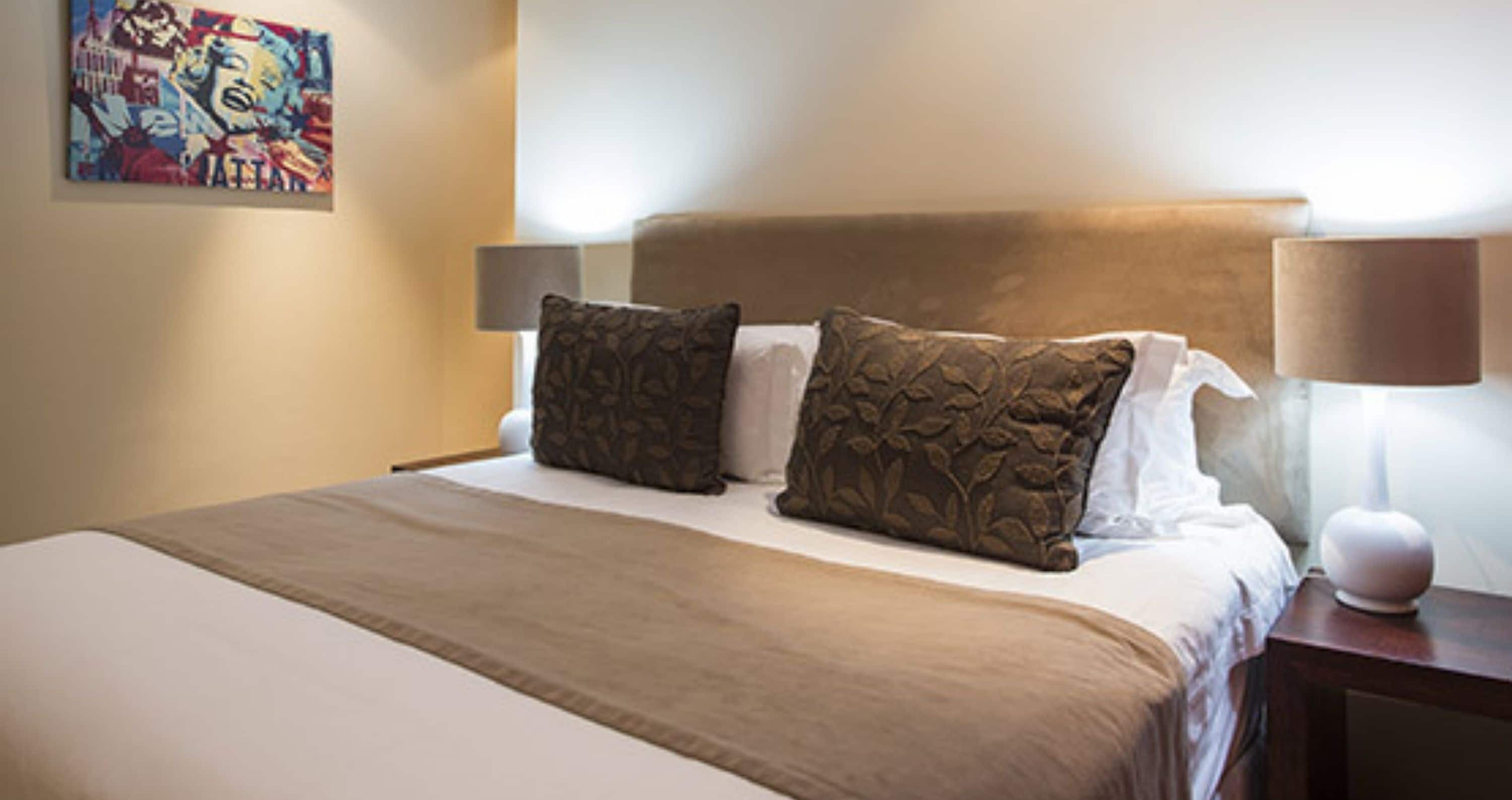 Cama bem arrumada com um cobre-leito marrom e duas almofadas decorativas, uma obra de arte na parede e um abajur sobre uma mesa de cabeceira de madeira.