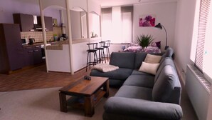 Uma sala de estar moderna com um sofá cinzento, uma mesa de centro de madeira e uma área de cozinha ao fundo.