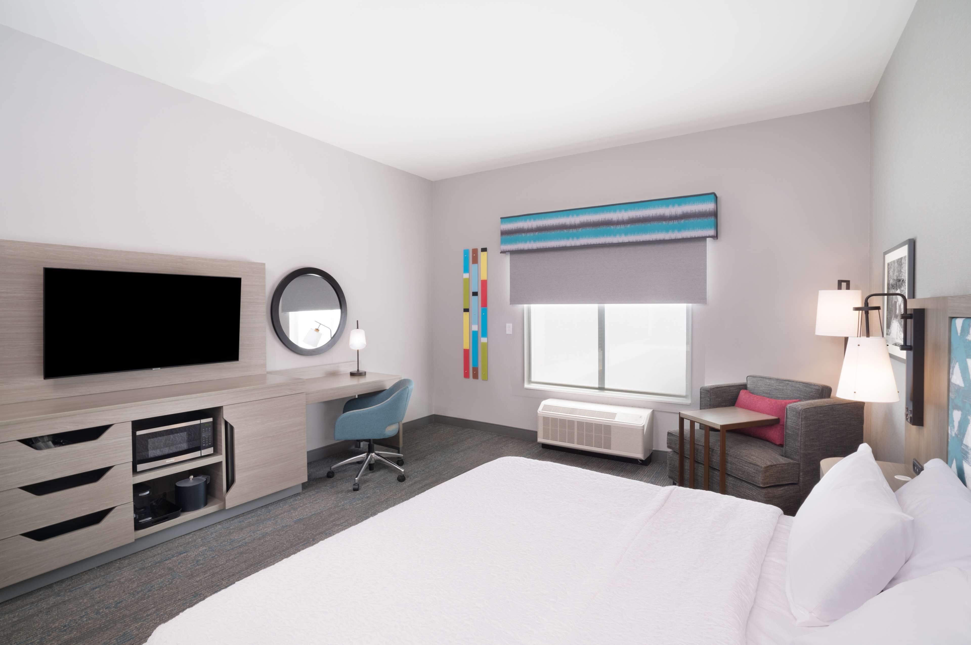 Quarto de hotel com cama, escrivaninha com cadeira, televisão, janela com vista e ar condicionado fixado na parede.
