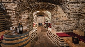 Uma adega de paredes de pedra com barris de madeira, uma estante para vinho e uma mesa coberta com um tecido vermelho.