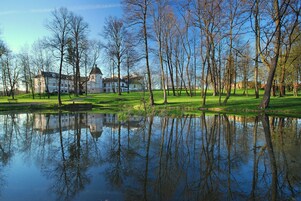 Um edifício grande refletido em águas calmas, rodeado por árvores e um espaço gramado.