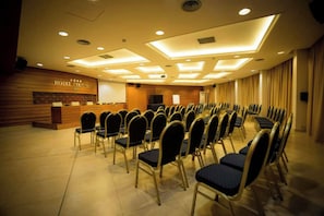 Uma sala de conferências de hotel com fileiras de cadeiras, uma mesa de recepção e uma tela grande.