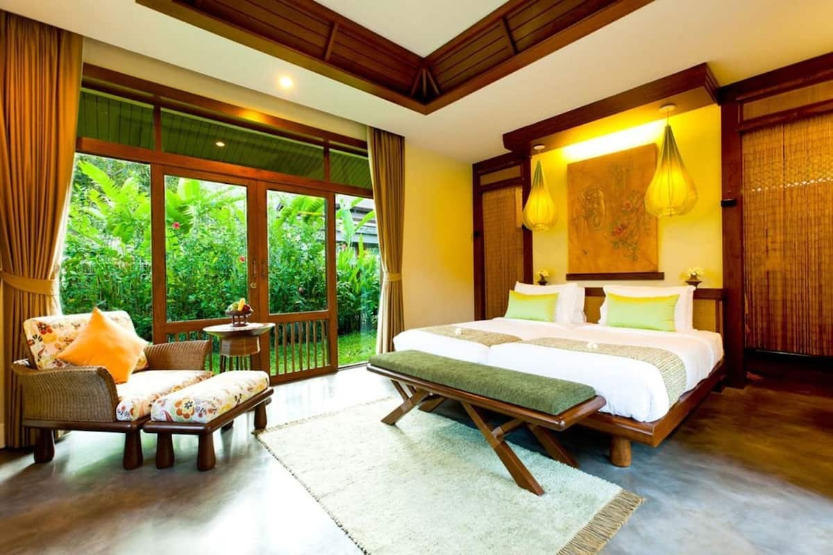 Quarto com uma cama grande, um espaço de estar com poltrona e mesa, e vista para vegetação exuberante através de grandes janelas.