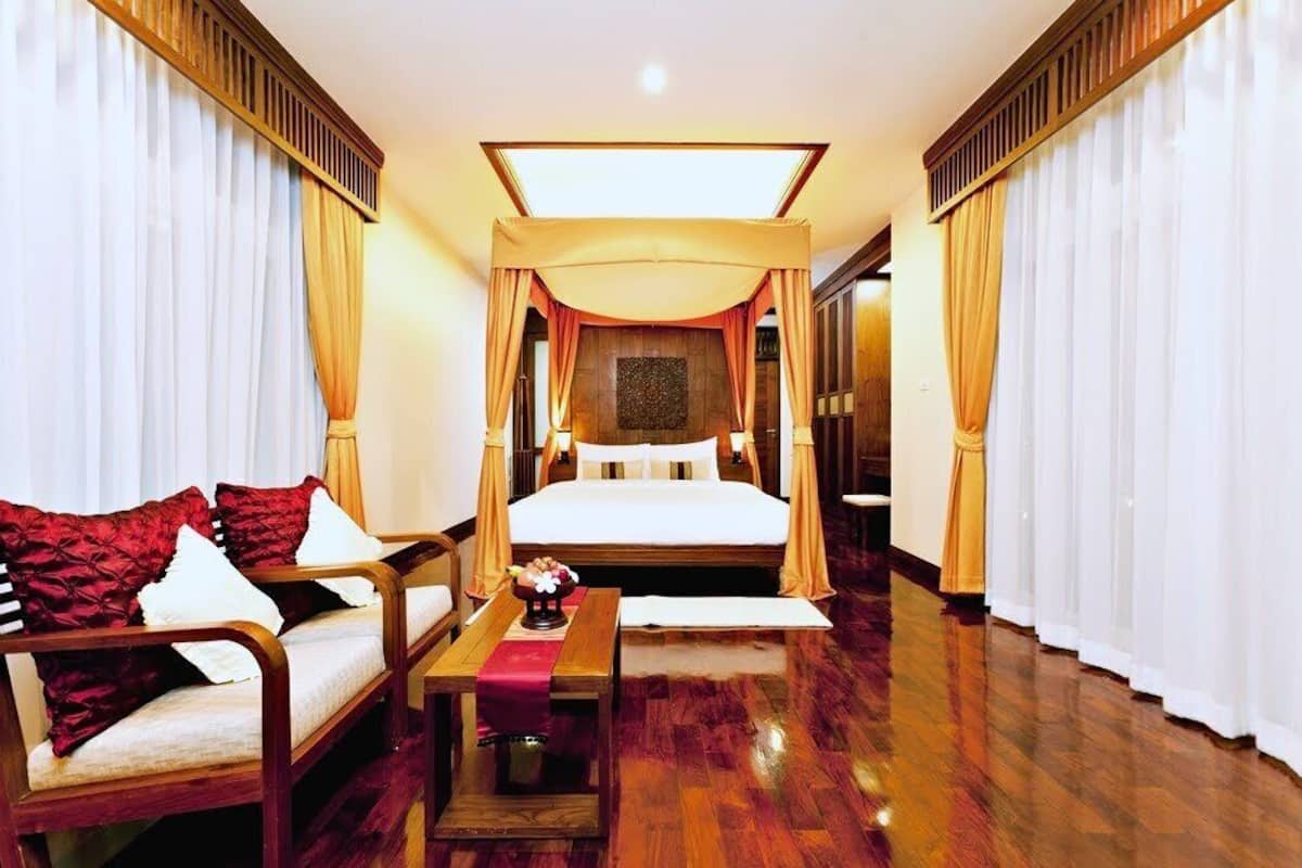 Quarto de hotel com uma cama, uma cadeira de madeira, uma mesinha e cortinas.