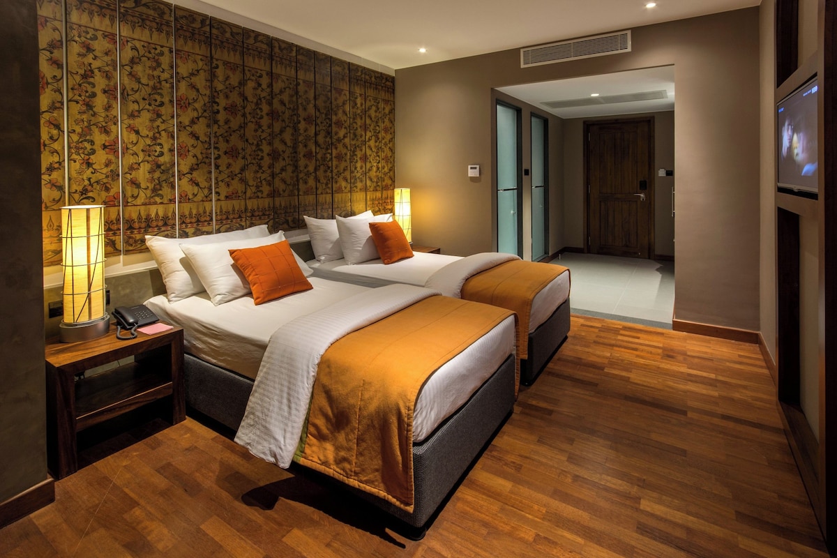 Quarto de hotel com duas camas, piso de madeira, uma janela grande e uma televisão na parede.