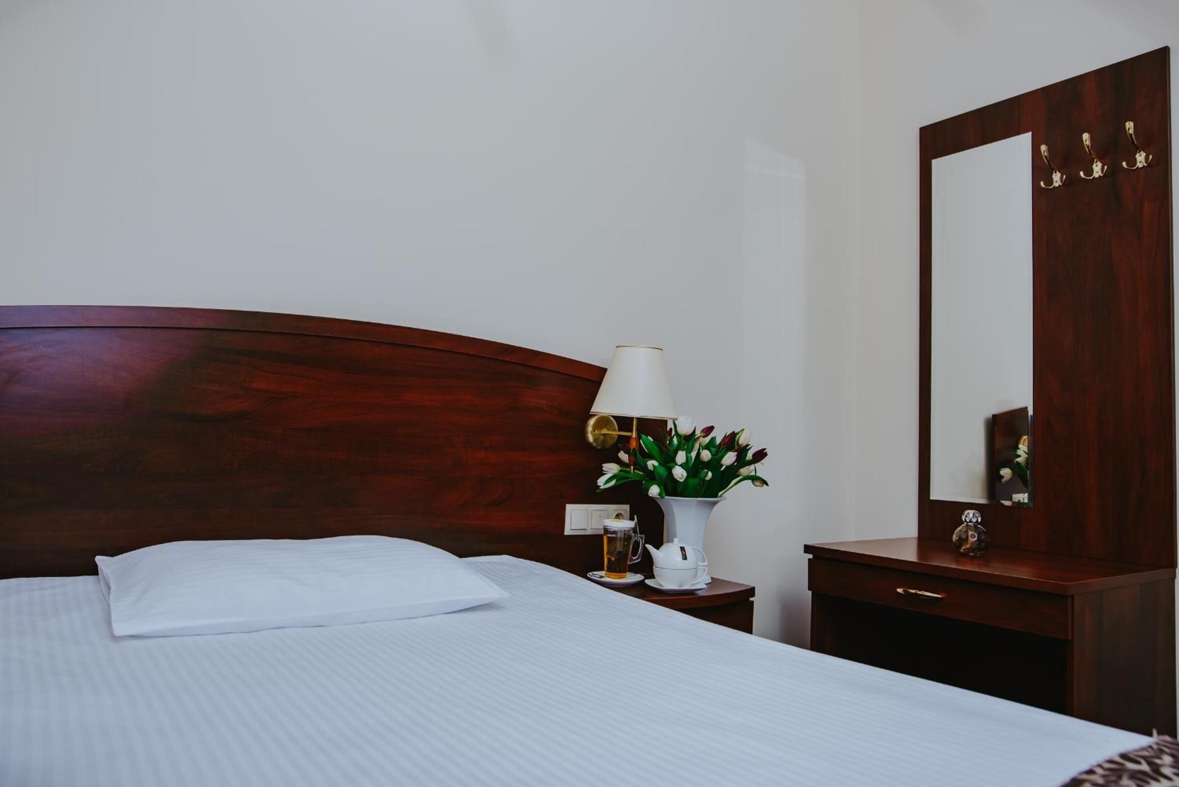 Cama bem arrumada com lençóis brancos, mesa de cabeceira com abajur, vaso com flores e espelho sobre cômoda de madeira.