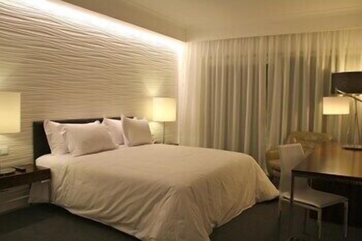 Quarto de hotel com uma cama grande, mesas de cabeceira, uma escrivaninha e uma cadeira.