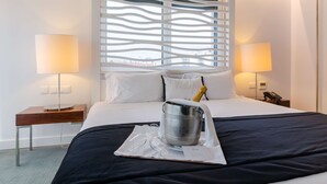 Quarto de hotel com uma cama, mesa de cabeceira com luminária, minibar e janela com um padrão ondulado peculiar.