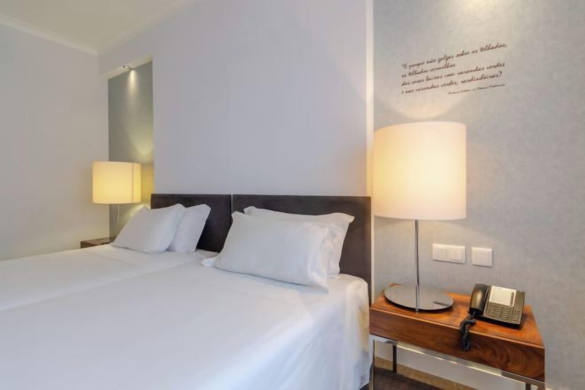 Quarto de hotel com uma cama bem arrumada, luminária de cabeceira, telefone e uma parede com uma citação.