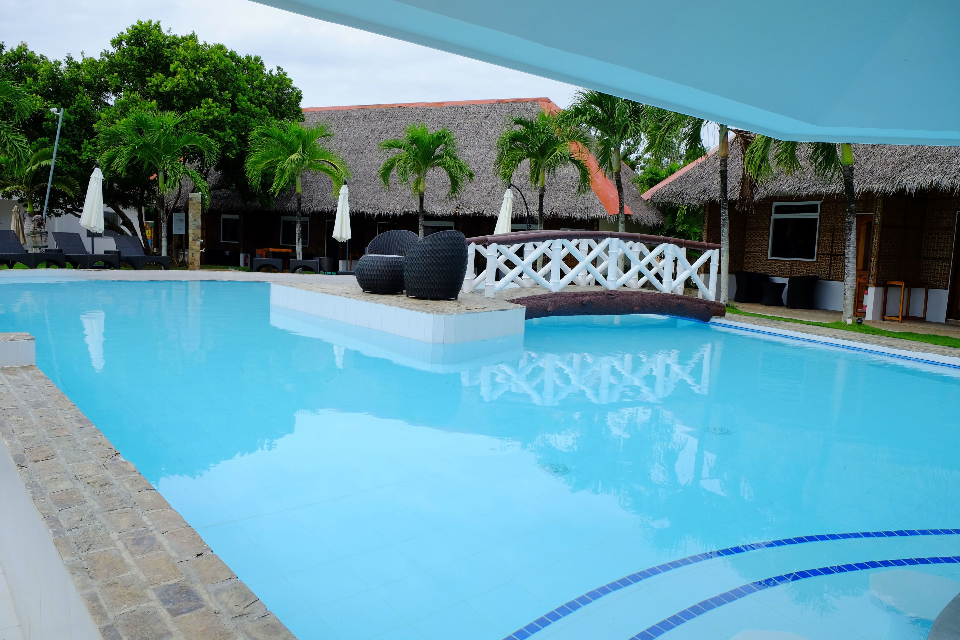 Piscina com espreguiçadeiras e guarda-sóis, cercada por edifícios com telhados de palha e palmeiras.